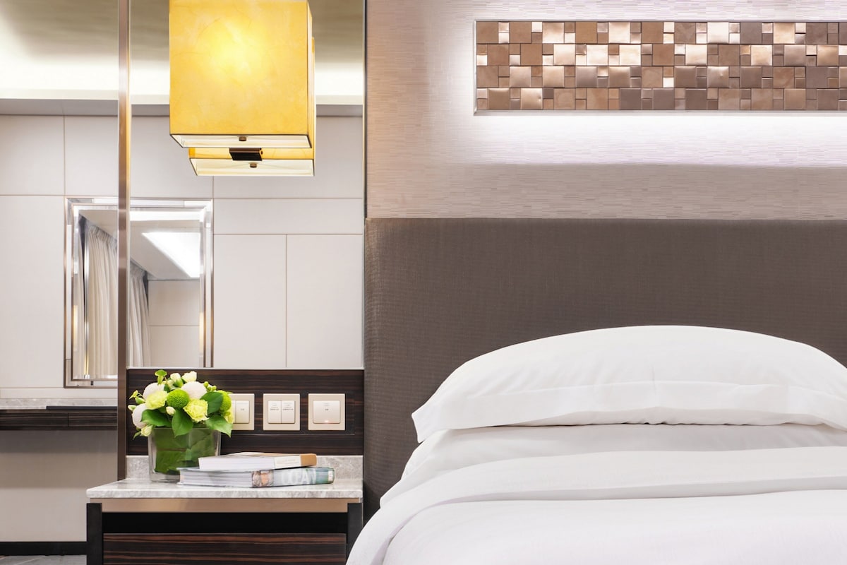 Quarto de hotel com uma cama, uma mesa de cabeceira com um vaso de flores, um espelho e uma luminária na parede.