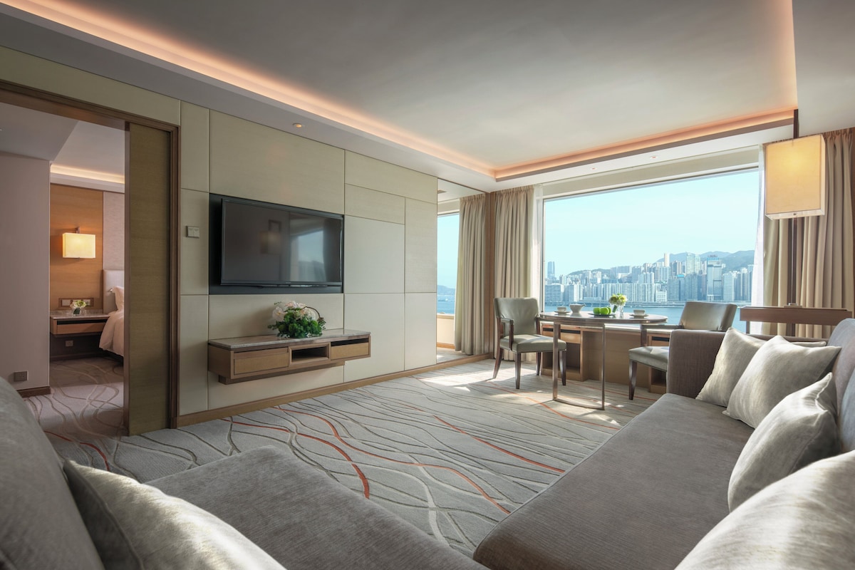 Quarto de hotel moderno com uma cama grande, televisão de tela plana na parede, área de jantar com mesa e cadeiras, e vista para a cidade pela janela.