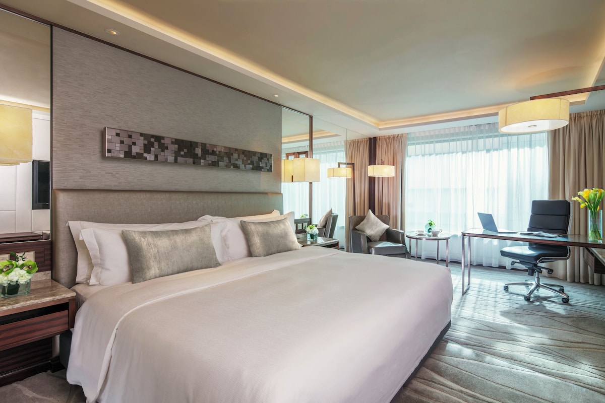 Quarto de hotel moderno com uma cama grande, uma escrivaninha com um laptop, uma cadeira e uma mesinha com um vaso de flores.