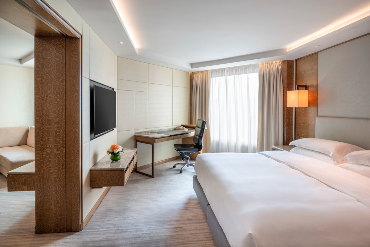 Quarto de hotel moderno com cama, escrivaninha, cadeira, televisão e janela com cortinas.