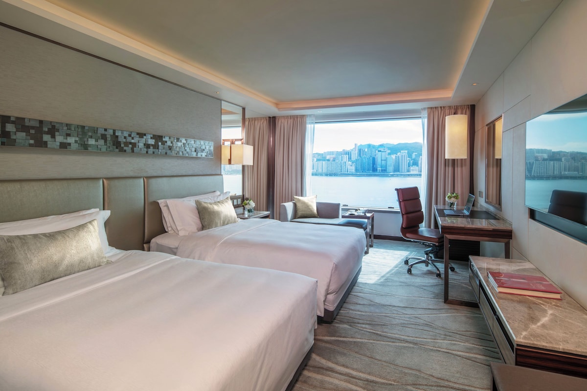 Quarto de hotel com duas camas, uma escrivaninha, uma cadeira e uma janela ampla com vista para a cidade.