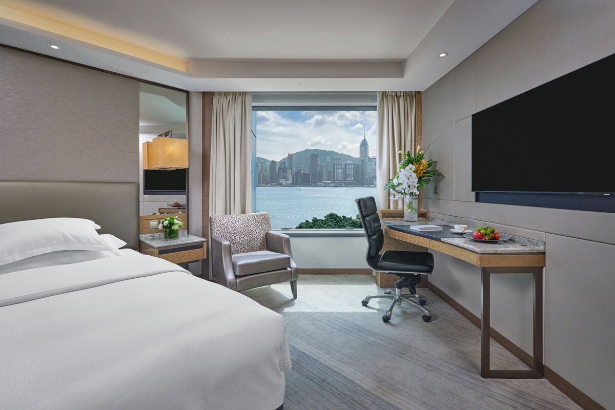 Quarto de hotel com cama, escrivaninha, poltrona, televisão e vista para a cidade e para a água.