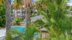 Uma área de piscina com palmeiras, uma estrutura com telhado de palha e um edifício com varandas.