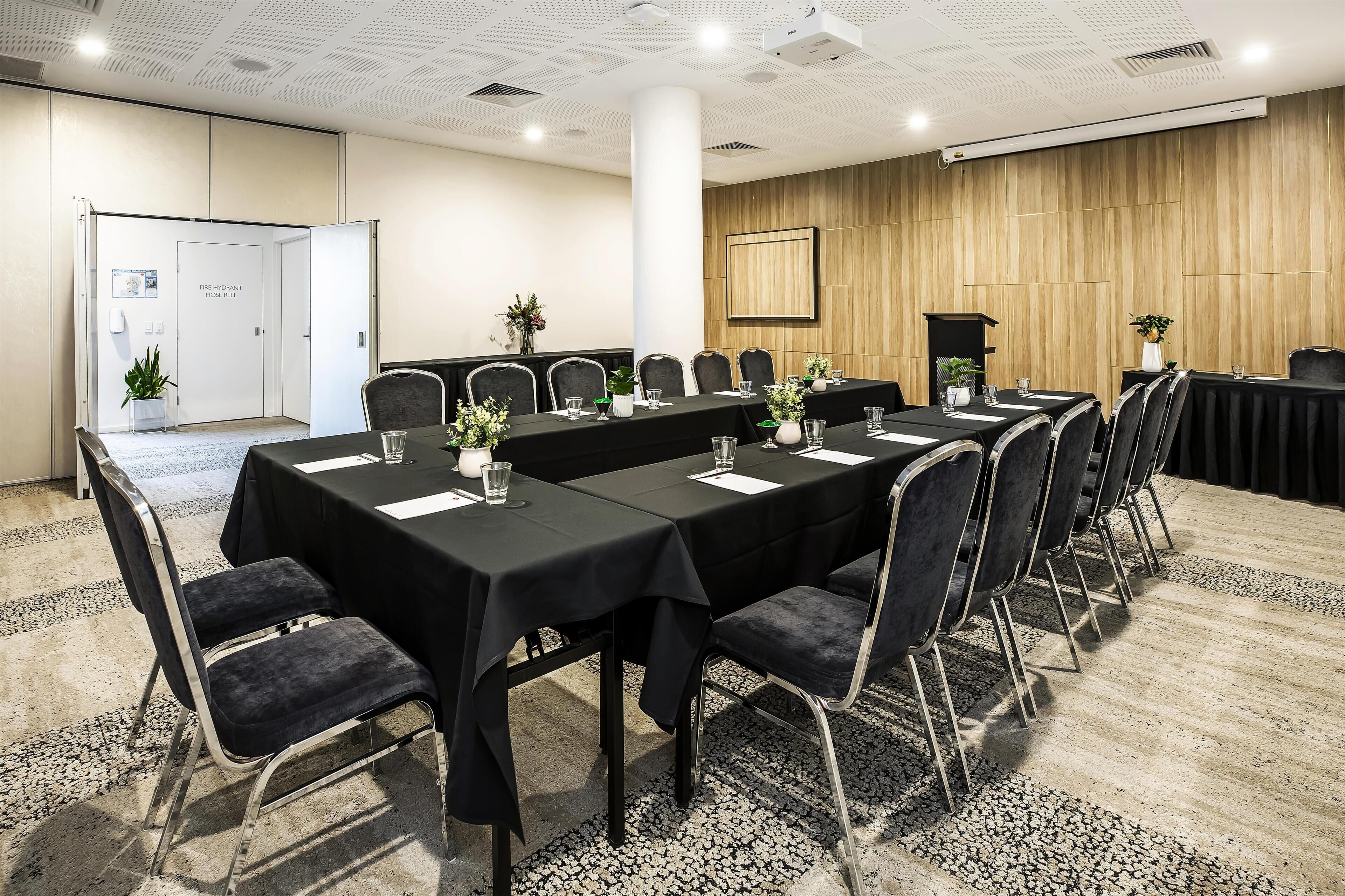Uma sala de conferências com uma mesa comprida, cadeiras, um pilar branco, um painel de parede de madeira e uma porta com a inscrição 'Fire Hydrant House Room'.