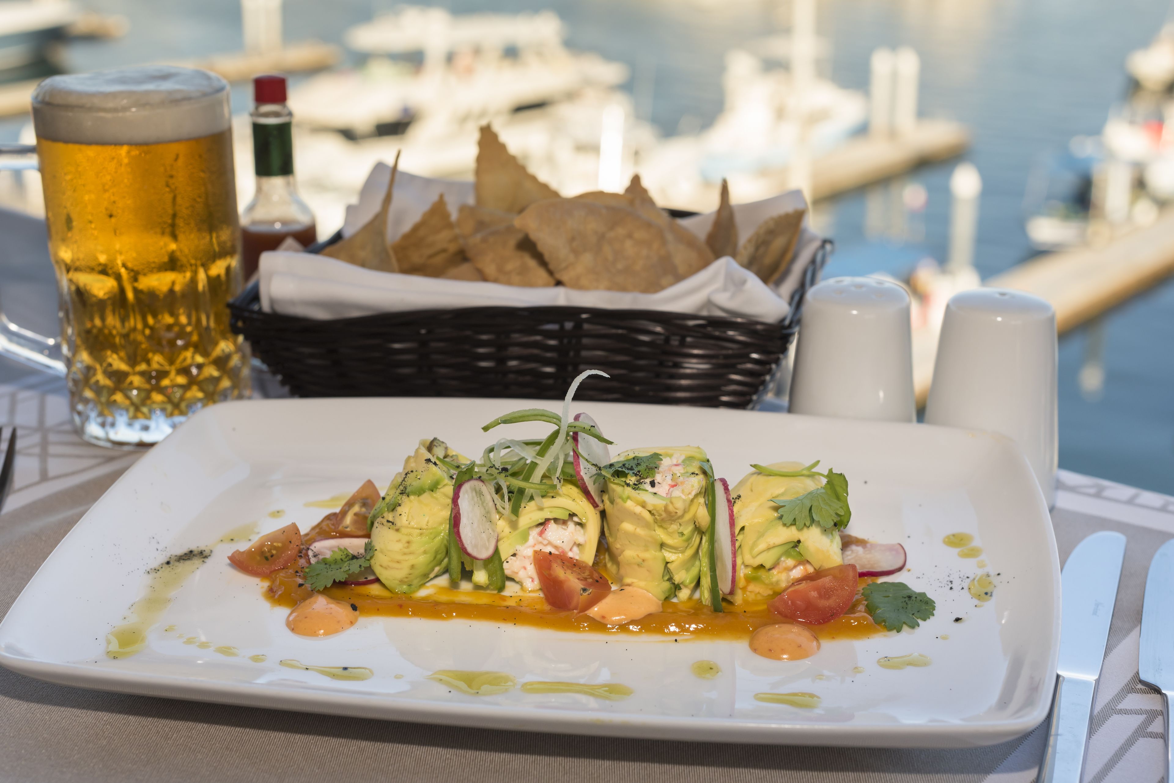 Prato de mariscos sobre um prato branco, um copo de cerveja e uma cesta de pão sobre uma mesa próxima a um porto de iates.