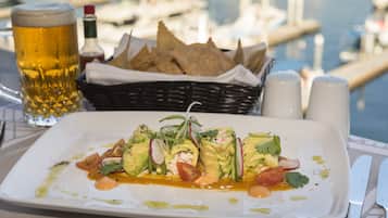 Prato de mariscos sobre um prato branco, um copo de cerveja e uma cesta de pão sobre uma mesa próxima a um porto de iates.