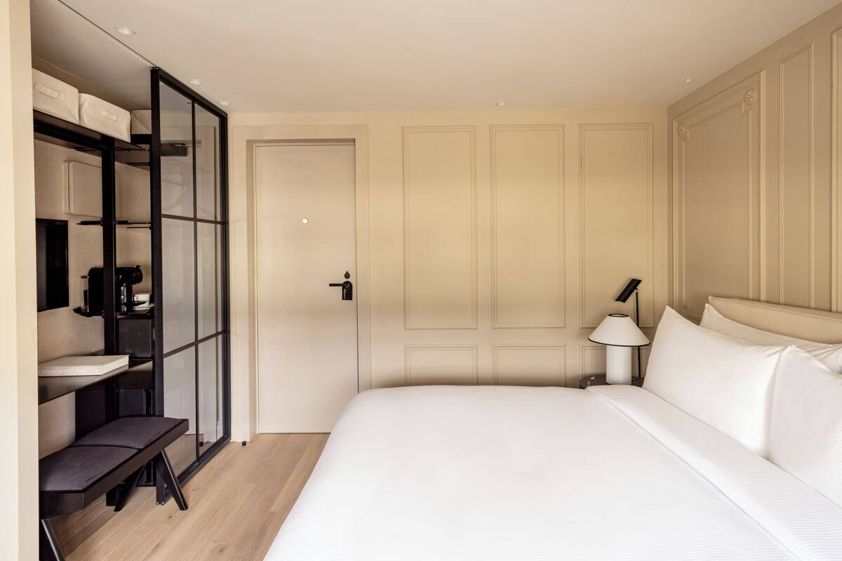 Quarto de hotel com uma cama, uma mesa com cadeira, uma luminária e um guarda-roupa.