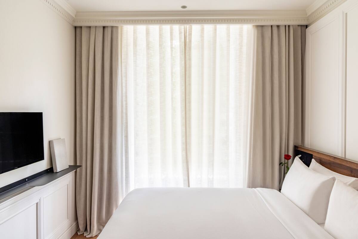 Quarto de hotel com uma cama grande, televisão de tela plana na parede e janela com cortinas leves.