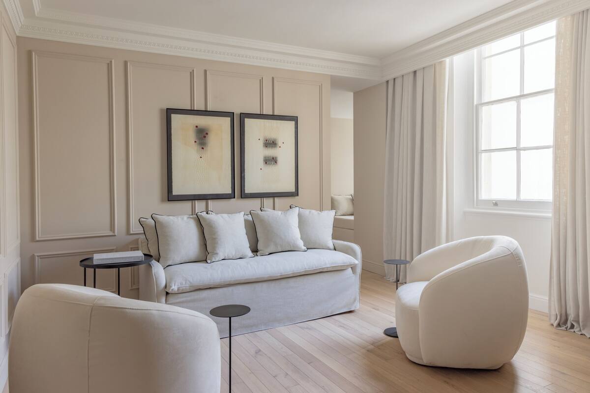 Uma sala de estar moderna com um sofá, duas poltronas e uma mesa de centro. Há duas obras de arte emolduradas na parede e uma janela com cortinas.