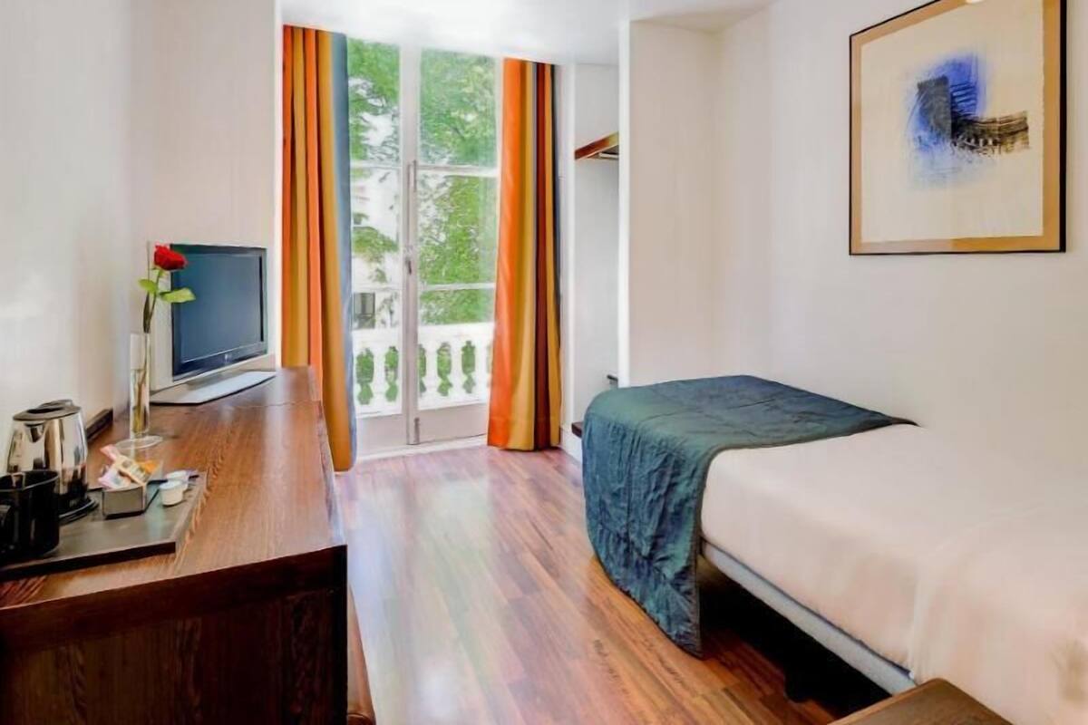 Quarto de hotel com cama, escrivaninha com computador, janela com cortinas e um quadro na parede.