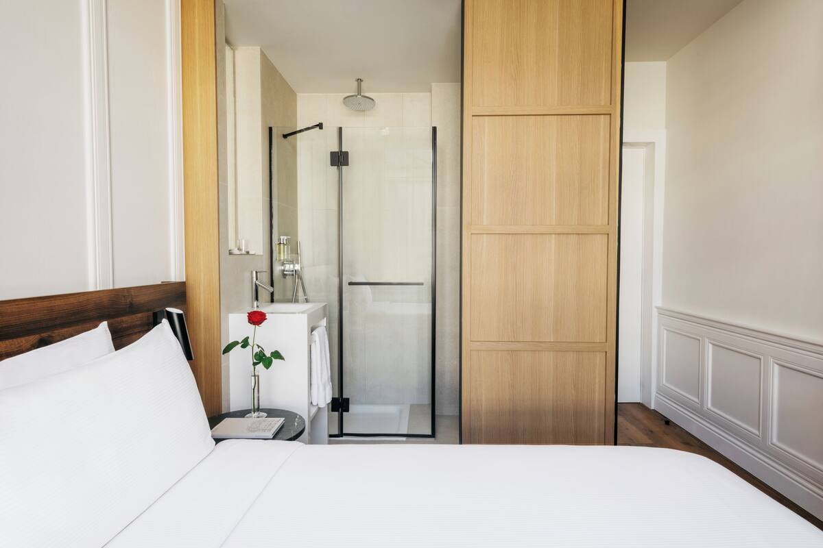 Quarto de hotel com cama, mesa de cabeceira, espelho, armário de madeira e chuveiro.