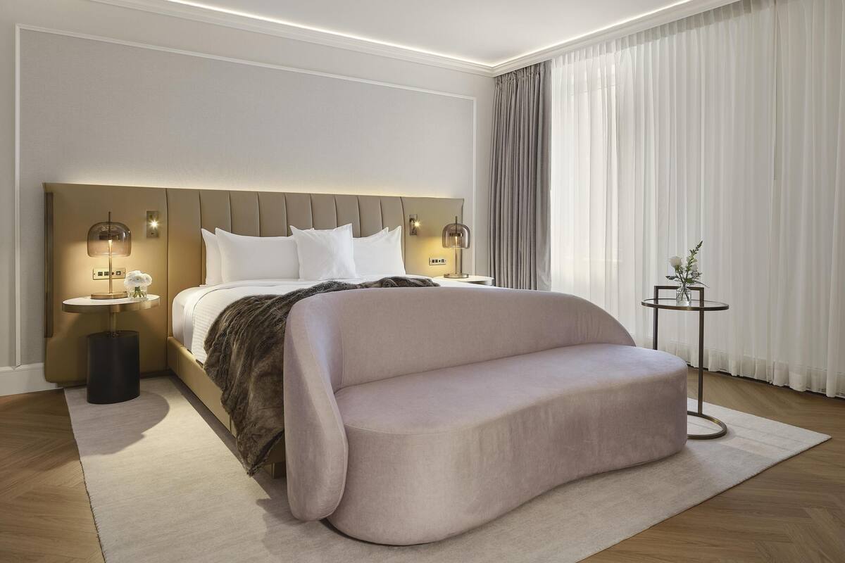 Quarto de hotel moderno com uma cama grande, duas mesas de cabeceira com luminárias, uma mesinha lateral e uma janela ampla com cortinas.
