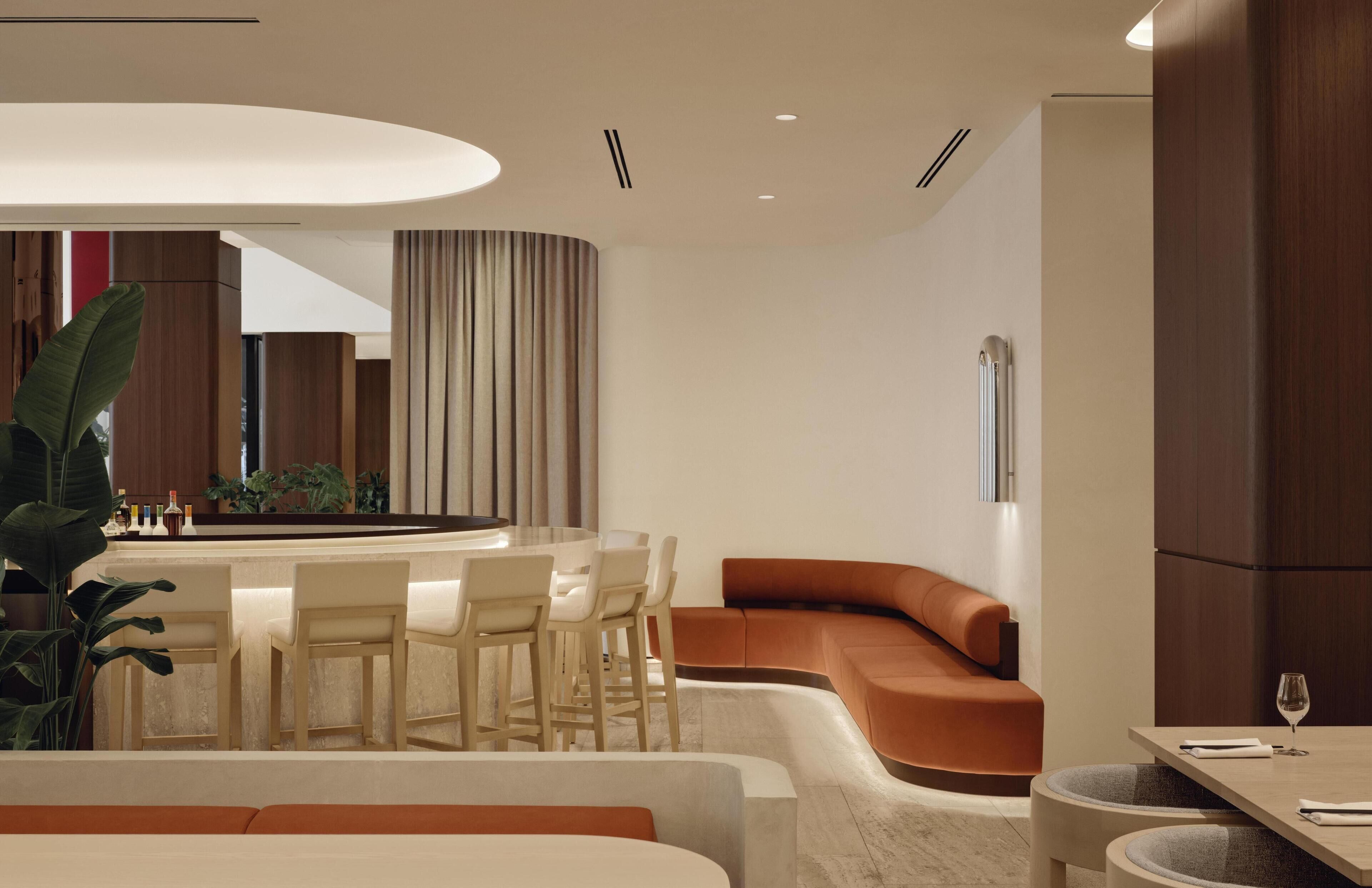 Uma área de jantar moderna com disposição de assentos em curva, uma ilha central com banquetas e uma grande planta em vaso.