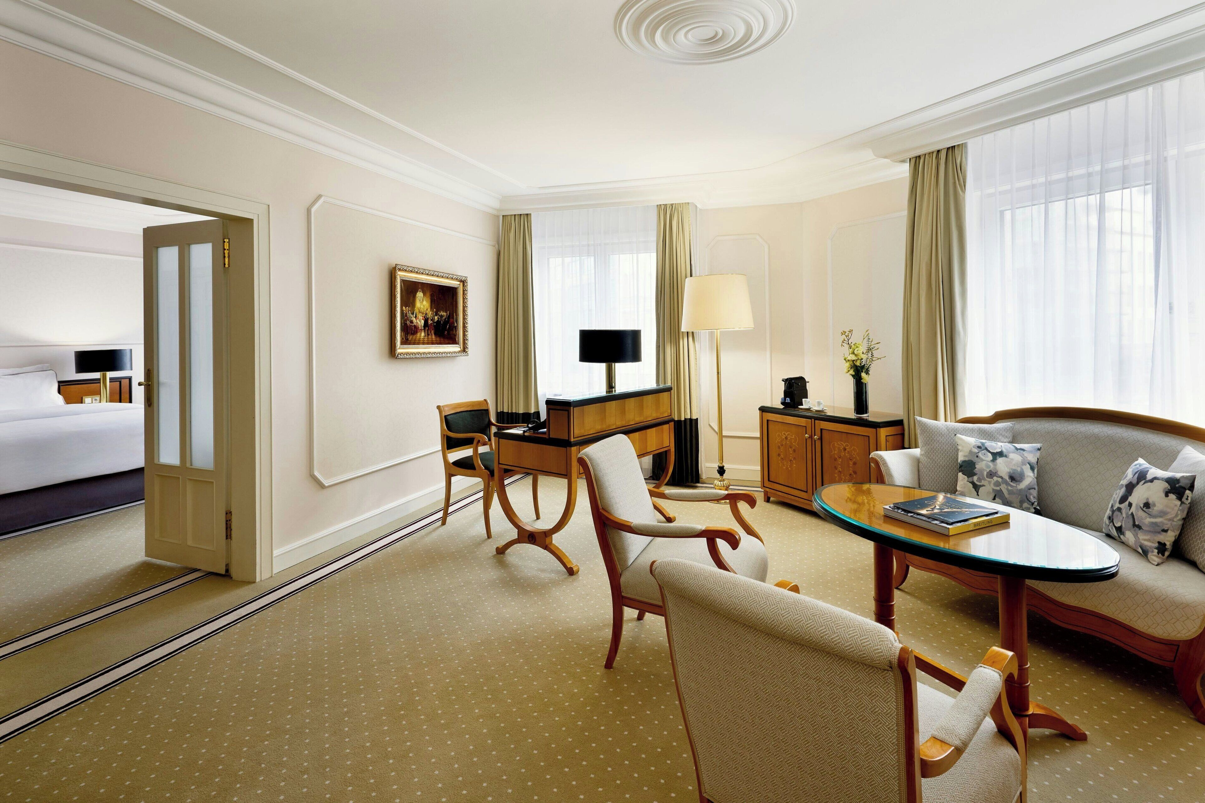 Quarto de hotel com cama, escrivaninha, sofá, mesa de centro e uma grande janela com cortinas.