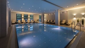 Piscina interna com piso revestido, rodeada por espreguiçadeiras e um espaço de estar com mesas.