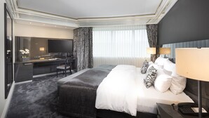 Quarto de hotel moderno com uma cama grande, mesa de cabeceira, luminária e uma escrivaninha com cadeira.