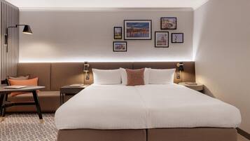 Quarto de hotel com uma cama grande, duas mesas de cabeceira, uma cadeira e uma escrivaninha. Há luminárias fixadas na parede e quadros emoldurados.