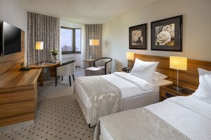 Quarto de hotel com duas camas, uma escrivaninha com cadeira, televisão e duas fotografias emolduradas na parede.