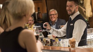 Ambiente de bar com um bartender servindo uma bebida. Há garrafas e copos no balcão.