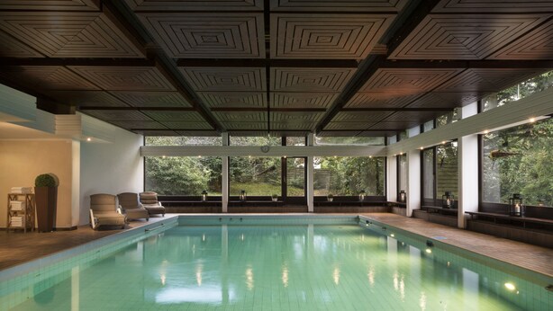 Uma grande piscina coberta com deck de madeira, espreguiçadeiras e amplas janelas com vista para o exterior.