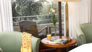 Quarto de hotel com varanda, uma mesa com abajur, um copo de suco de laranja, um vaso com flores e uma cadeira.