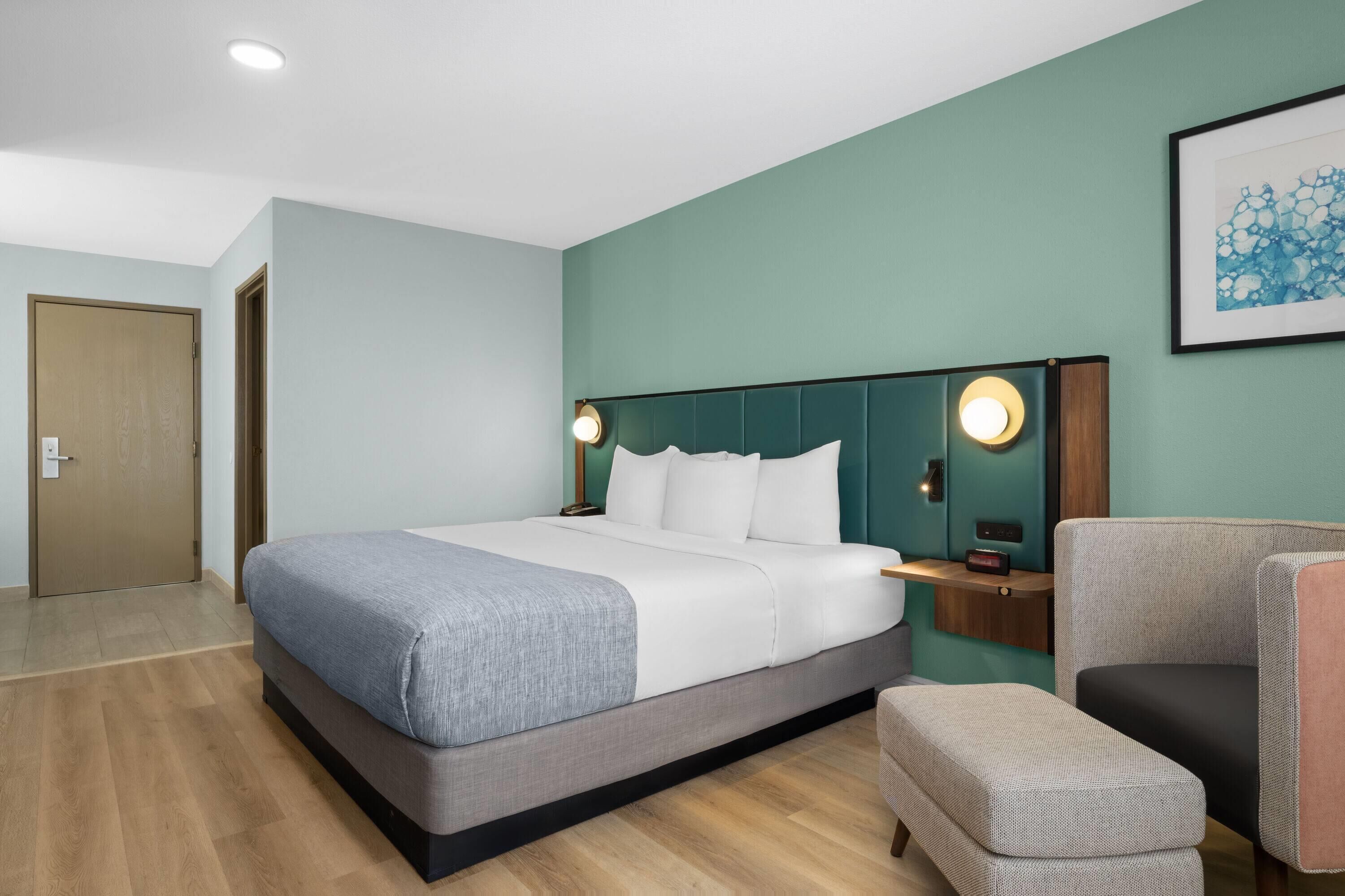 Quarto de hotel com cama, mesa de cabeceira, cadeira e luminária fixada na parede.