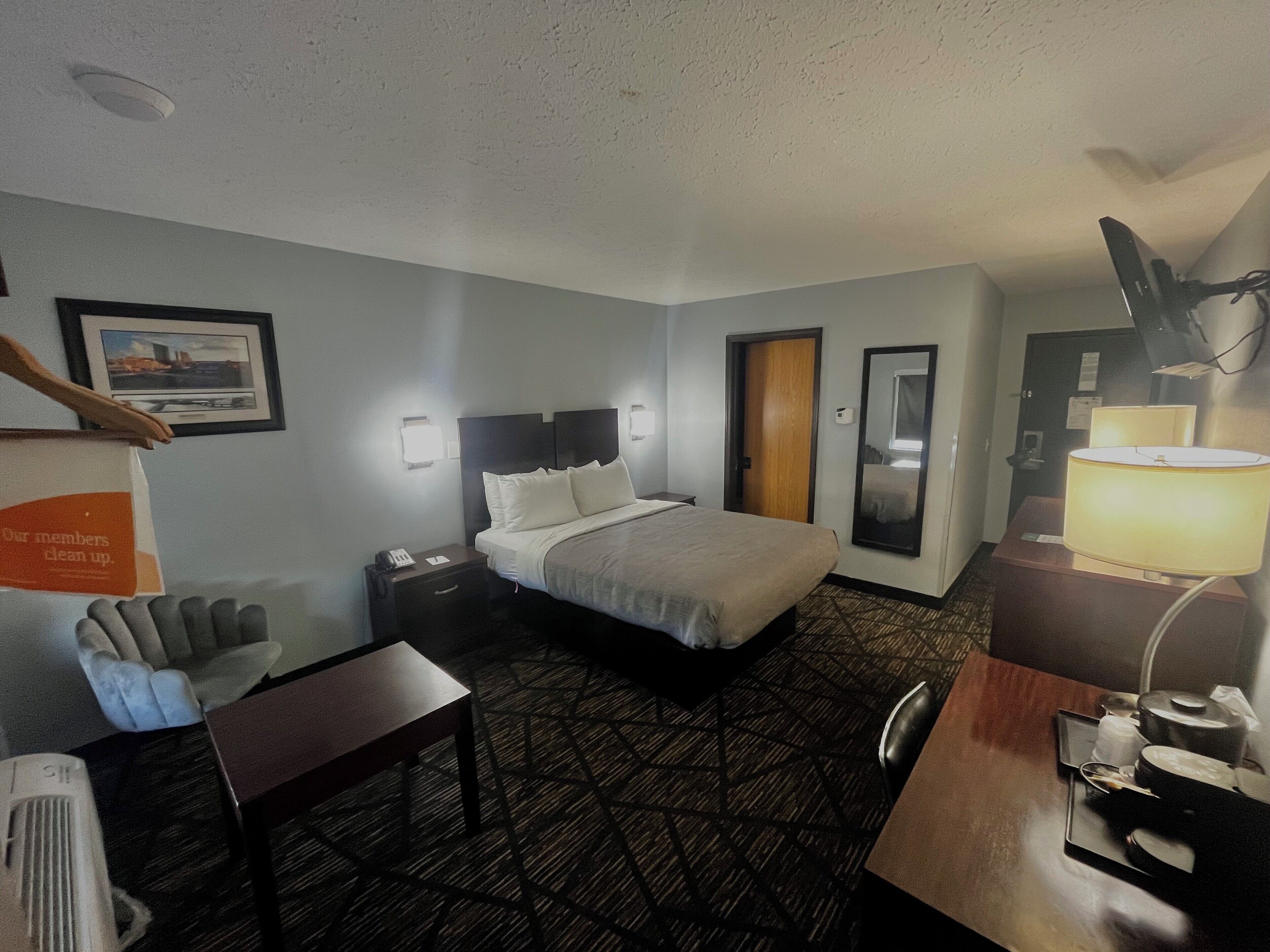 Quarto de hotel com cama, mesa de cabeceira, cadeira, escrivaninha, abajur e um quadro na parede.