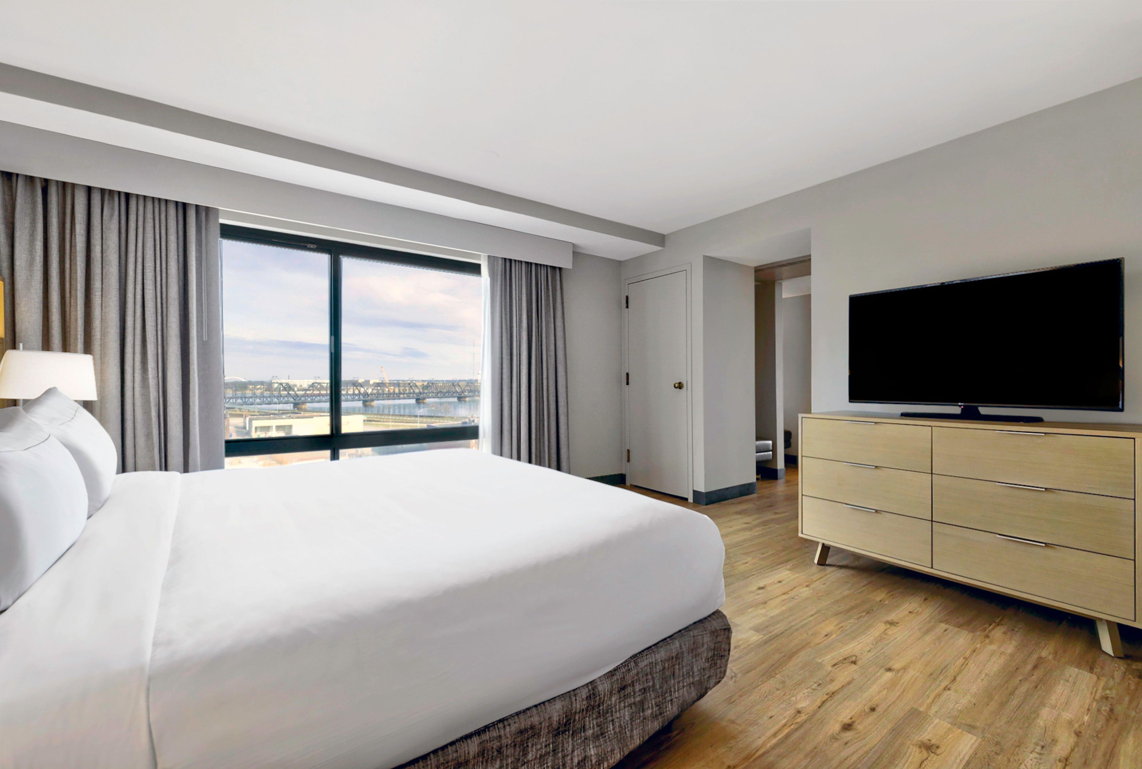 Quarto de hotel com uma cama grande, televisão, cômoda e vista para a paisagem urbana através de uma janela ampla.