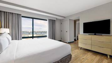 Quarto de hotel com uma cama grande, televisão, cômoda e vista para a paisagem urbana através de uma janela ampla.