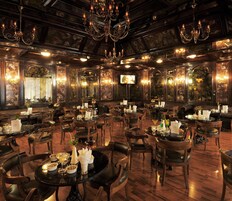 Uma área de jantar luxuosa, com várias mesas e cadeiras, lustres ornamentados e um espelho grande refletindo a decoração do espaço.
