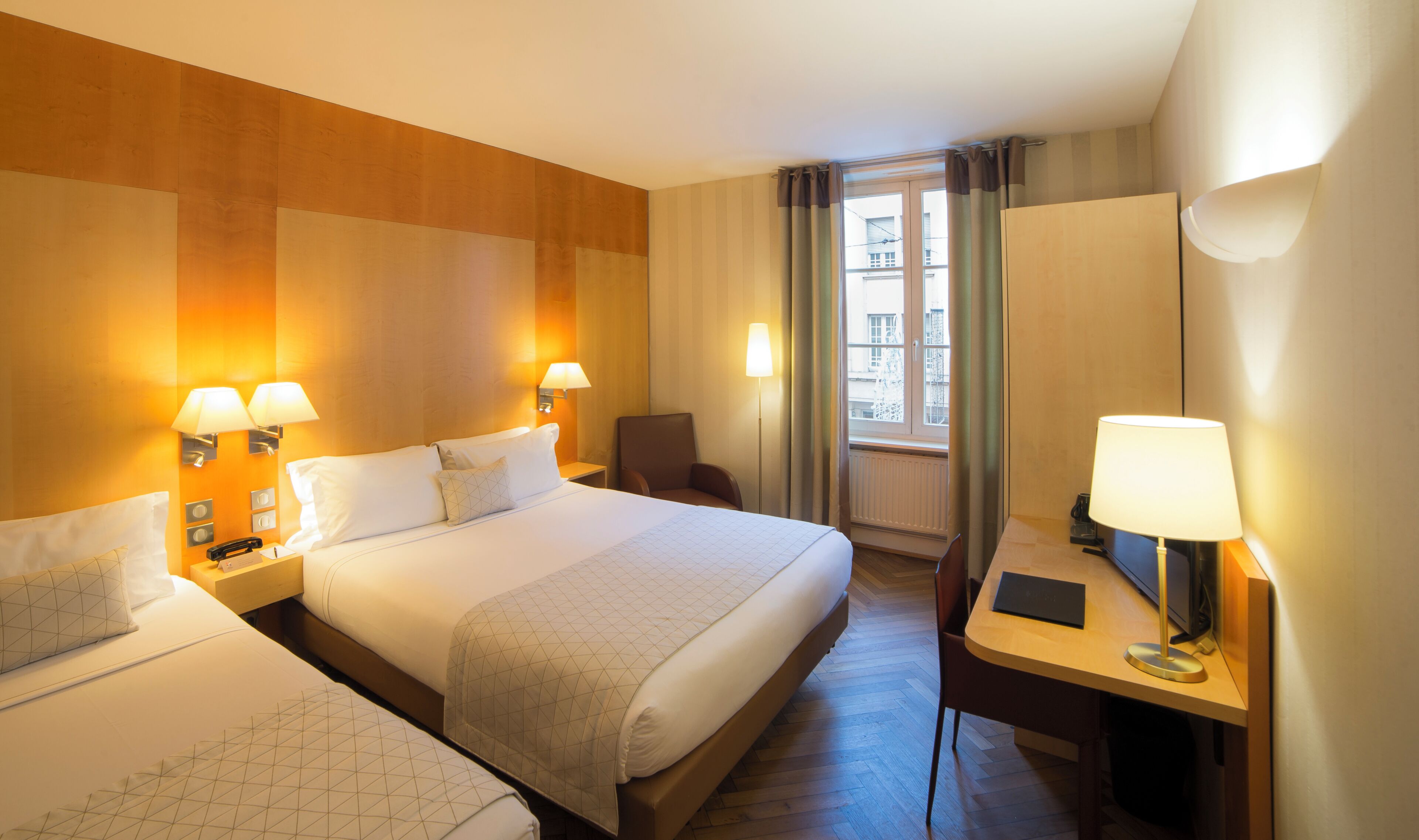 Quarto de hotel com duas camas, uma escrivaninha, uma cadeira, um abajur e uma janela com cortinas.