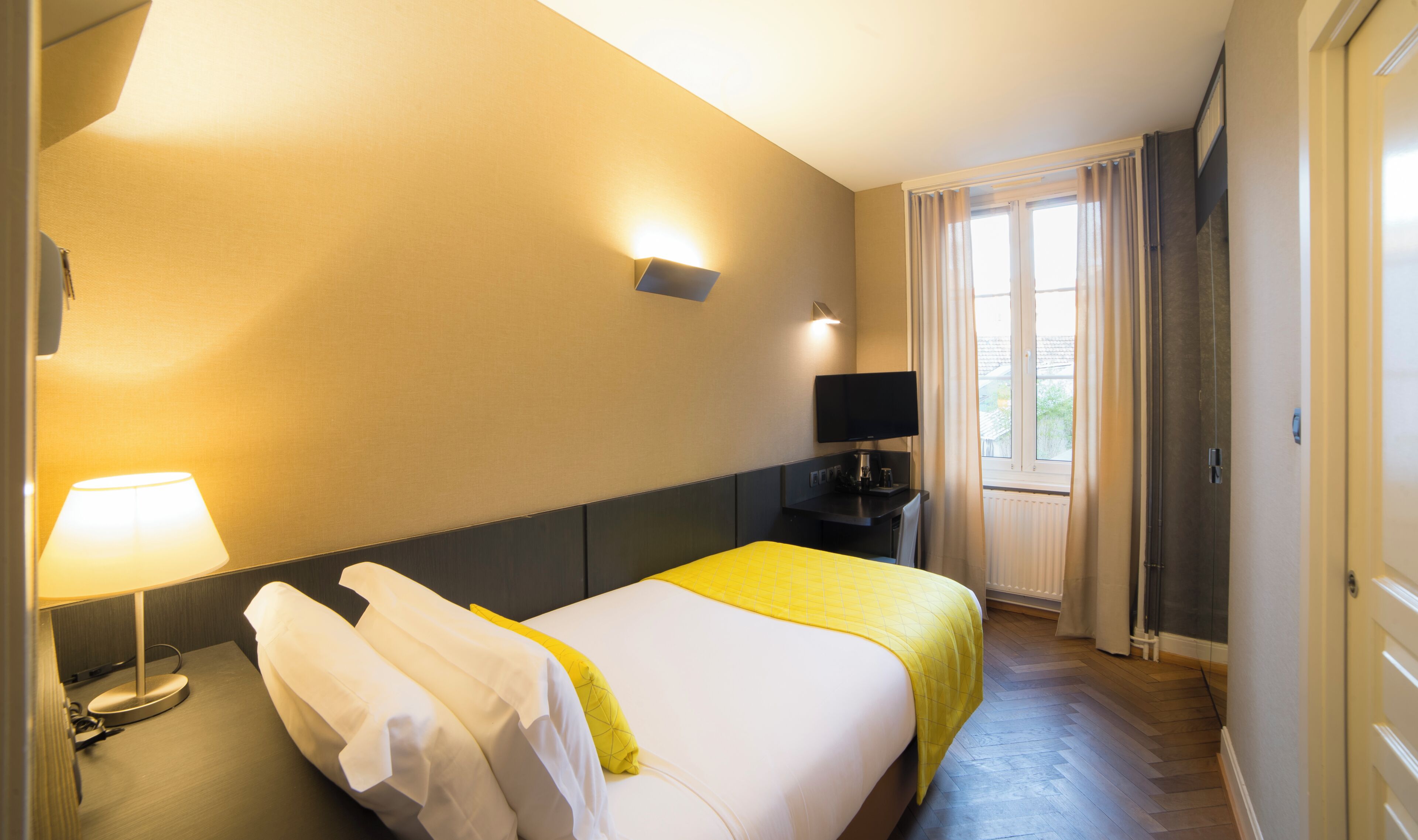 Quarto de hotel com cama, escrivaninha com computador, luminária e janela com cortinas.