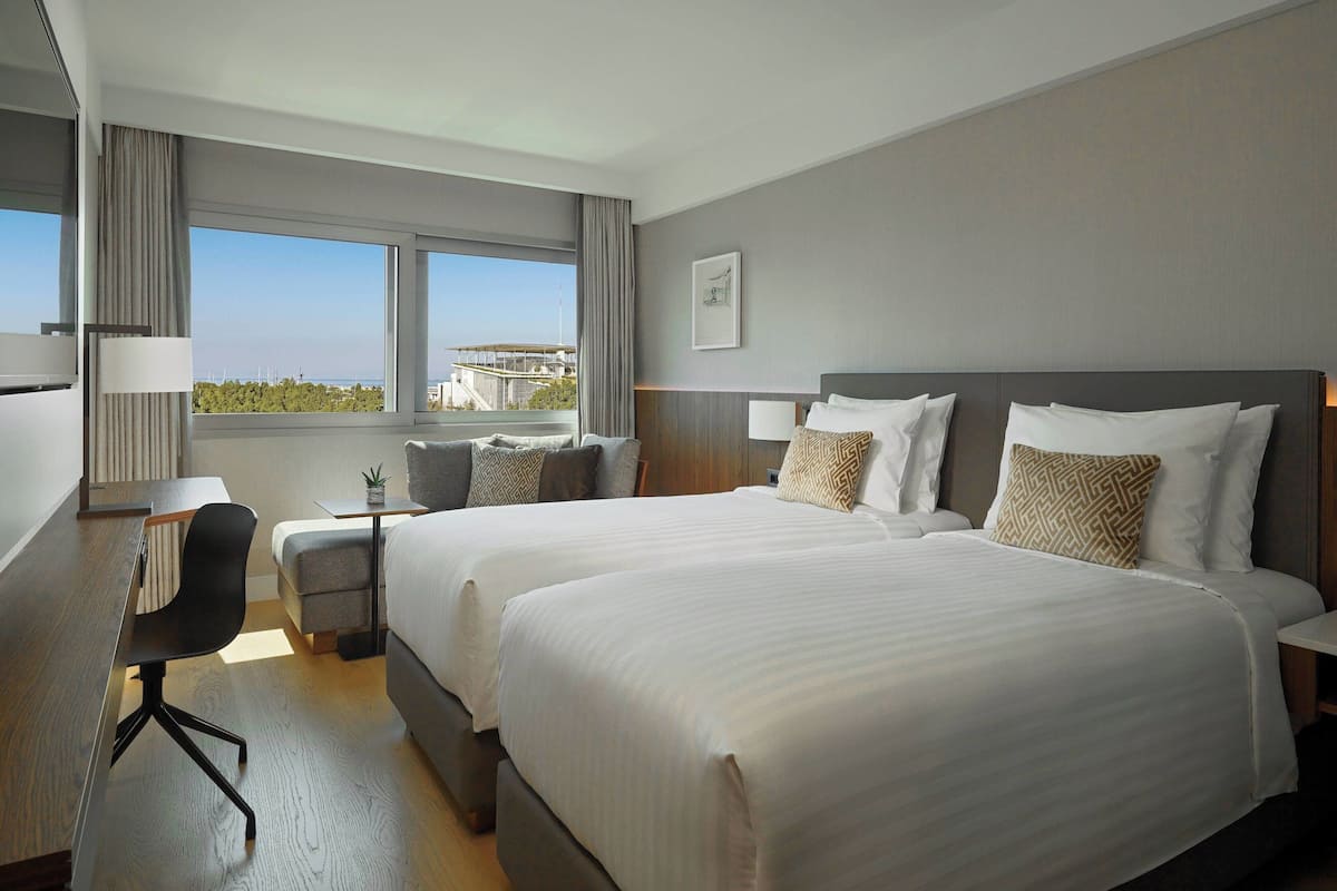 Quarto de hotel com duas camas, uma escrivaninha, uma cadeira e vista para a paisagem urbana.