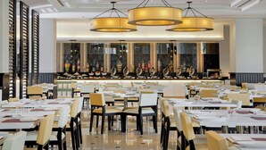 Uma sala de jantar espaçosa, com várias mesas e cadeiras, um bar central e um lustre moderno.