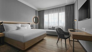 Quarto de hotel moderno com uma cama grande, uma escrivaninha, uma cadeira, uma TV e vista para a cidade através de amplas janelas.
