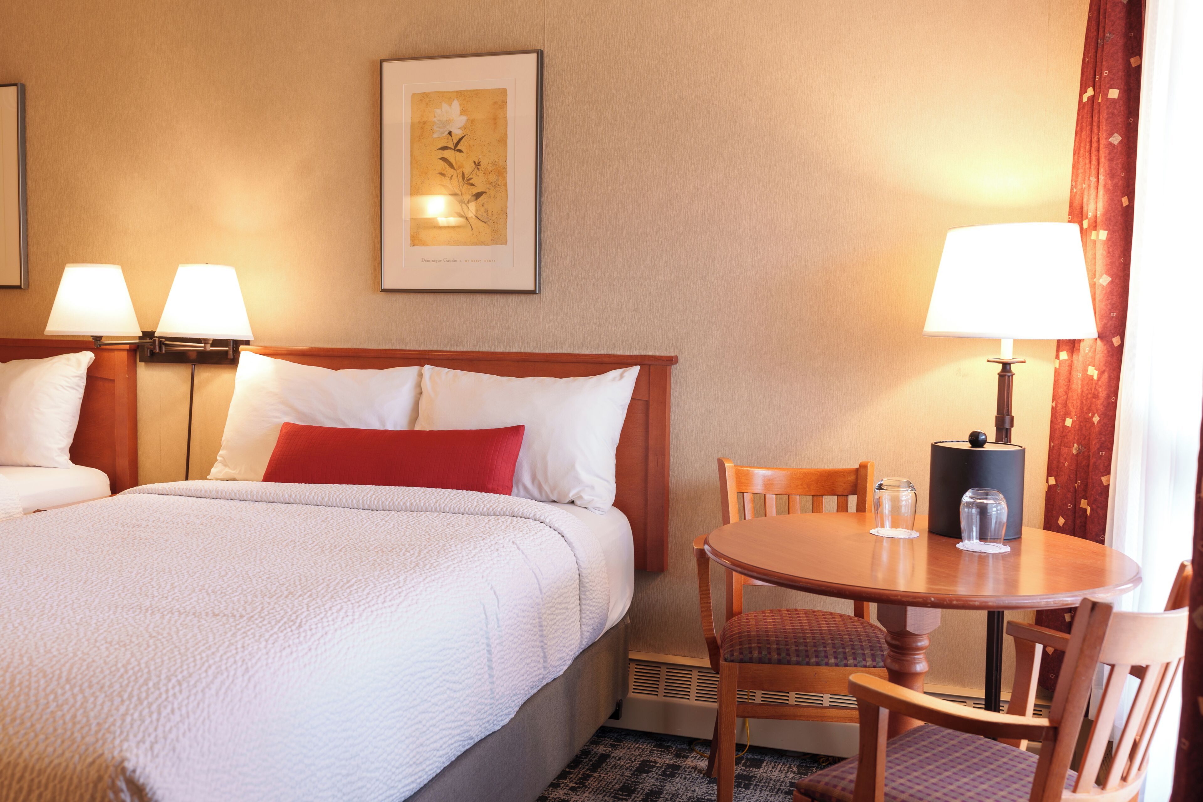 Quarto de hotel com uma cama, duas luminárias de cabeceira, um quadro na parede, uma mesa de jantar com cadeiras e uma janela com cortinas.