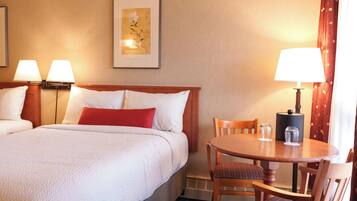 Quarto de hotel com uma cama, duas luminárias de cabeceira, um quadro na parede, uma mesa de jantar com cadeiras e uma janela com cortinas.