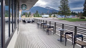 Um deck de madeira com mesas e cadeiras, com vista para uma estrada com veículos e montanhas ao fundo.