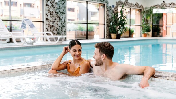Um casal relaxando em uma banheira de hidromassagem com vista para a área da piscina e paisagem externa.