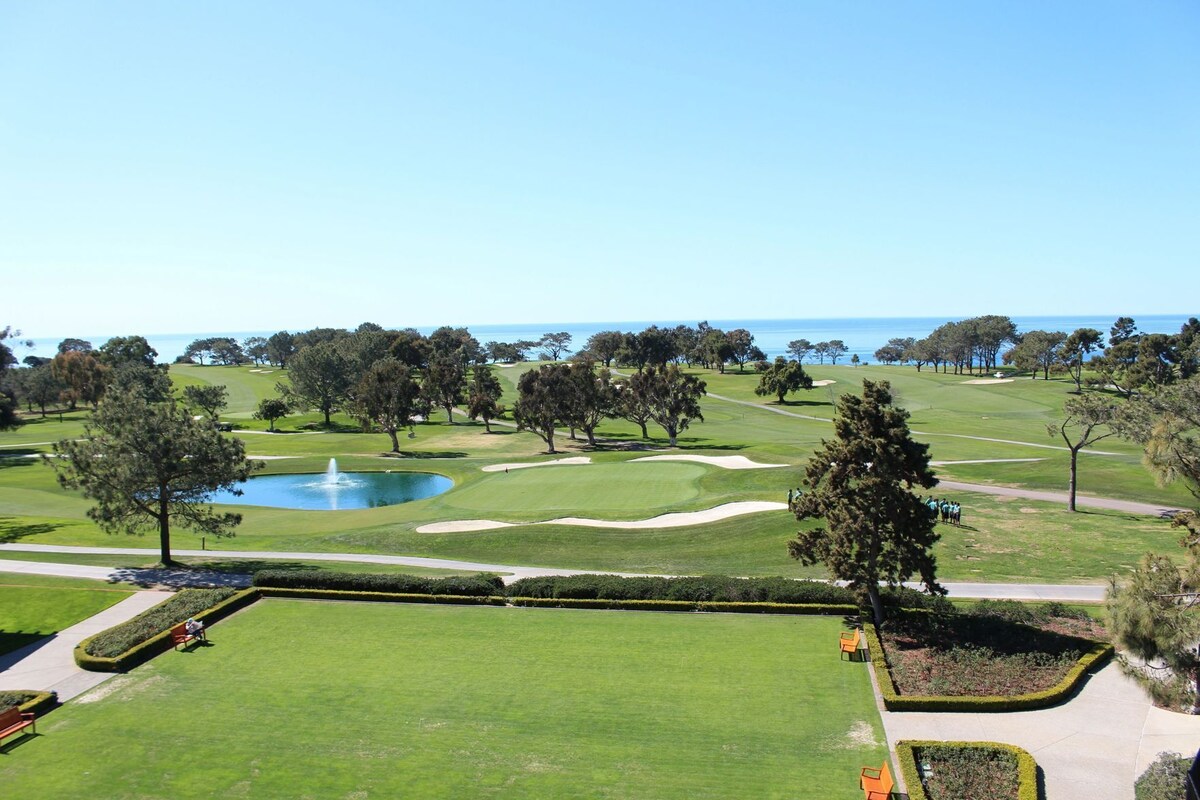Um campo de golfe com lago, árvores e vista para o oceano.