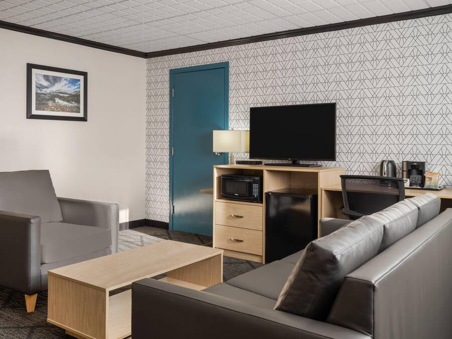 Quarto de hotel moderno com sofá, poltrona, TV, micro-ondas e escrivaninha com luminária.