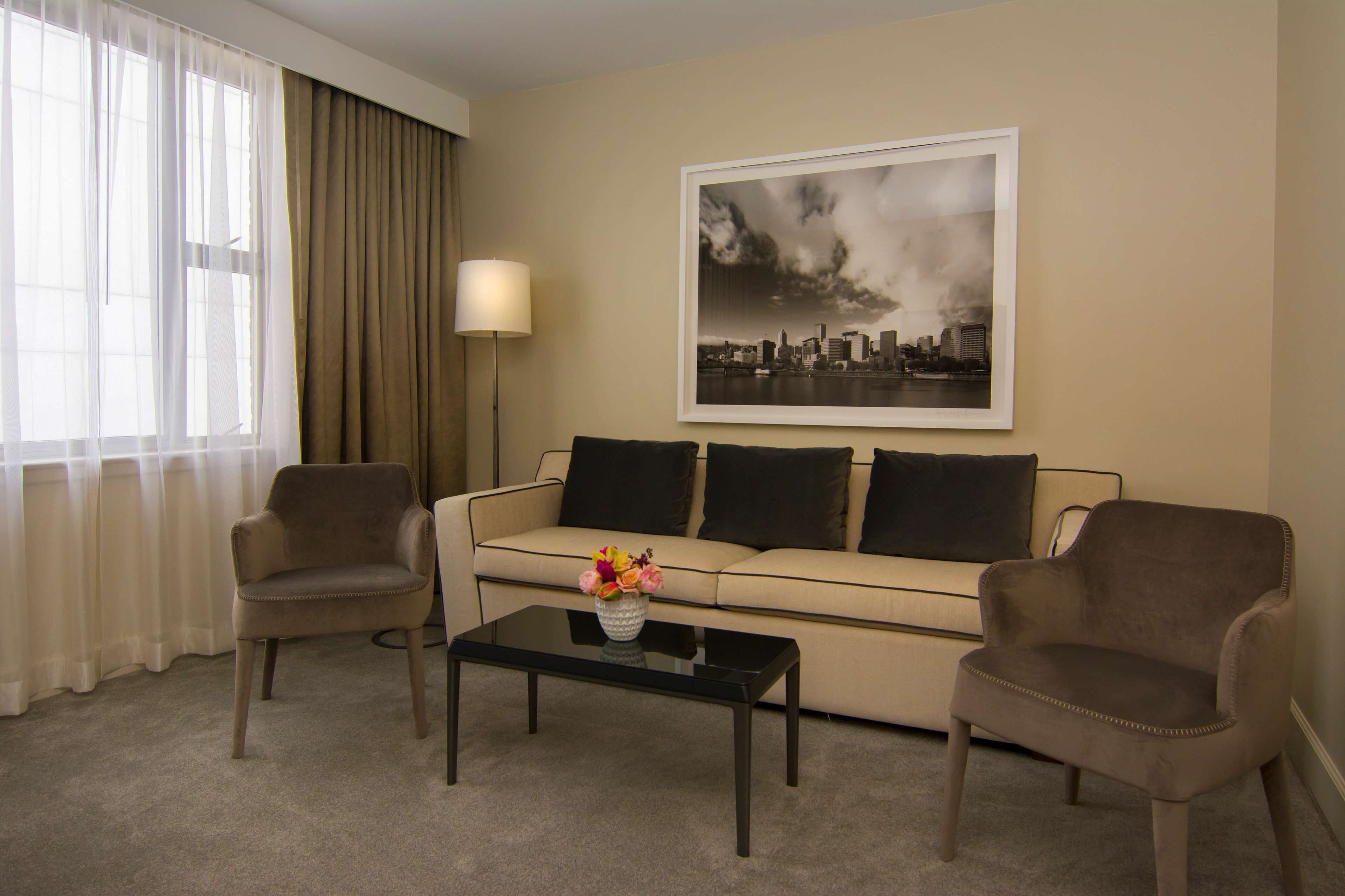 Quarto de hotel com sofá bege, duas poltronas, uma mesa de centro com um vaso de flores, um abajur e uma pintura de paisagem urbana na parede.
