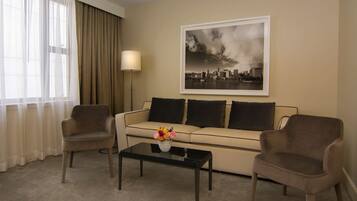 Quarto de hotel com sofá bege, duas poltronas, uma mesa de centro com um vaso de flores, um abajur e uma pintura de paisagem urbana na parede.
