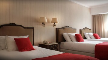 Quarto de hotel com duas camas, cada uma com uma almofada vermelha decorativa, uma mesa de cabeceira com telefone e luminárias fixadas na parede.