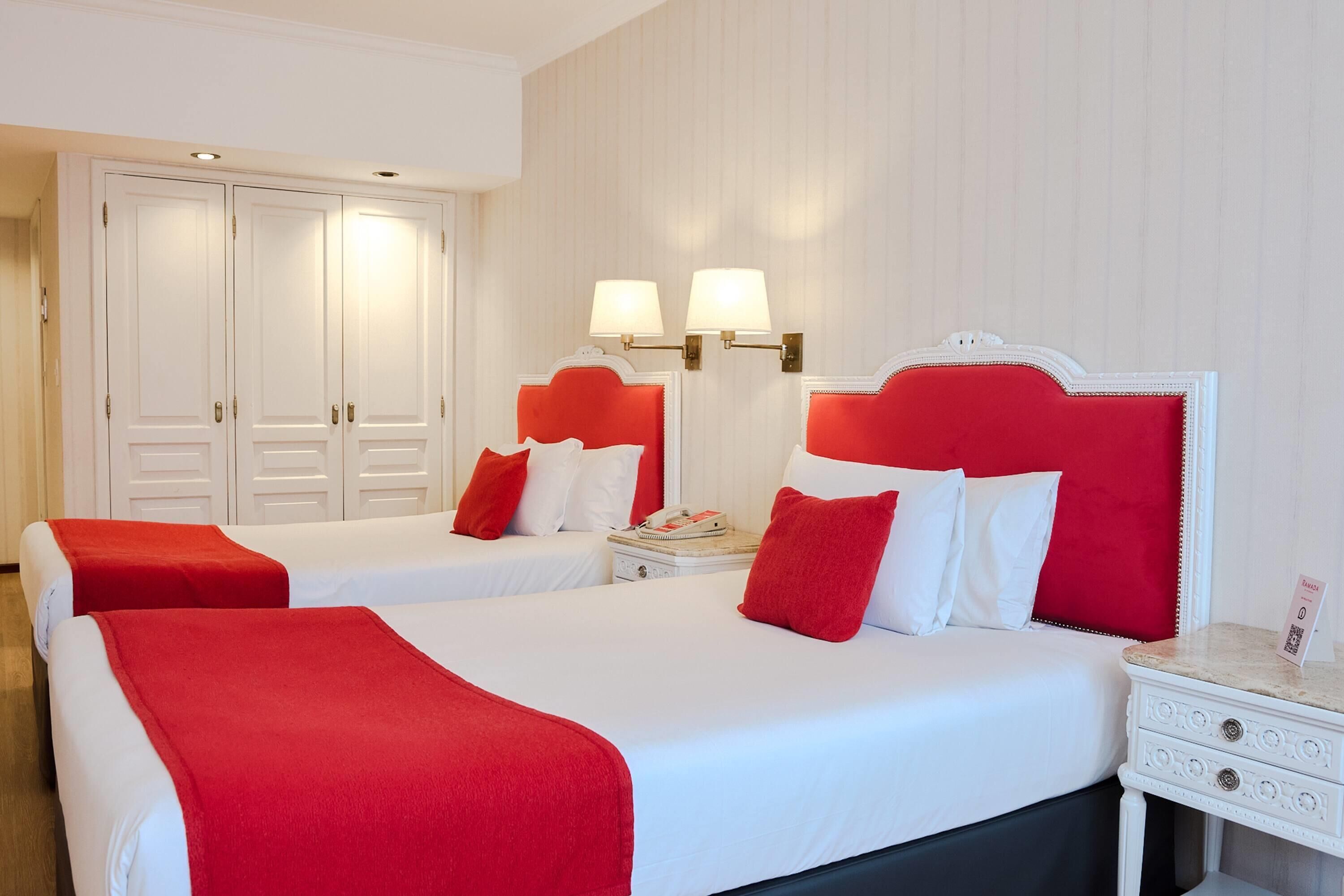 Quarto de hotel com duas camas, roupa de cama branca, detalhes em vermelho, uma mesa de cabeceira com luminária e um guarda-roupa.
