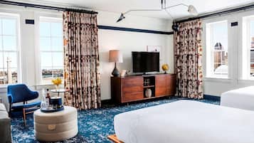 Quarto de hotel com uma cama grande, uma poltrona azul, cômoda de madeira com televisão e cortinas florais.