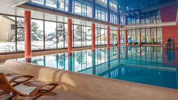 Piscina interna com paredes de vidro transparente, espreguiçadeiras de madeira e vista para árvores cobertas de neve lá fora.