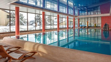 Piscina interna com paredes de vidro transparente, espreguiçadeiras de madeira e vista para árvores cobertas de neve lá fora.