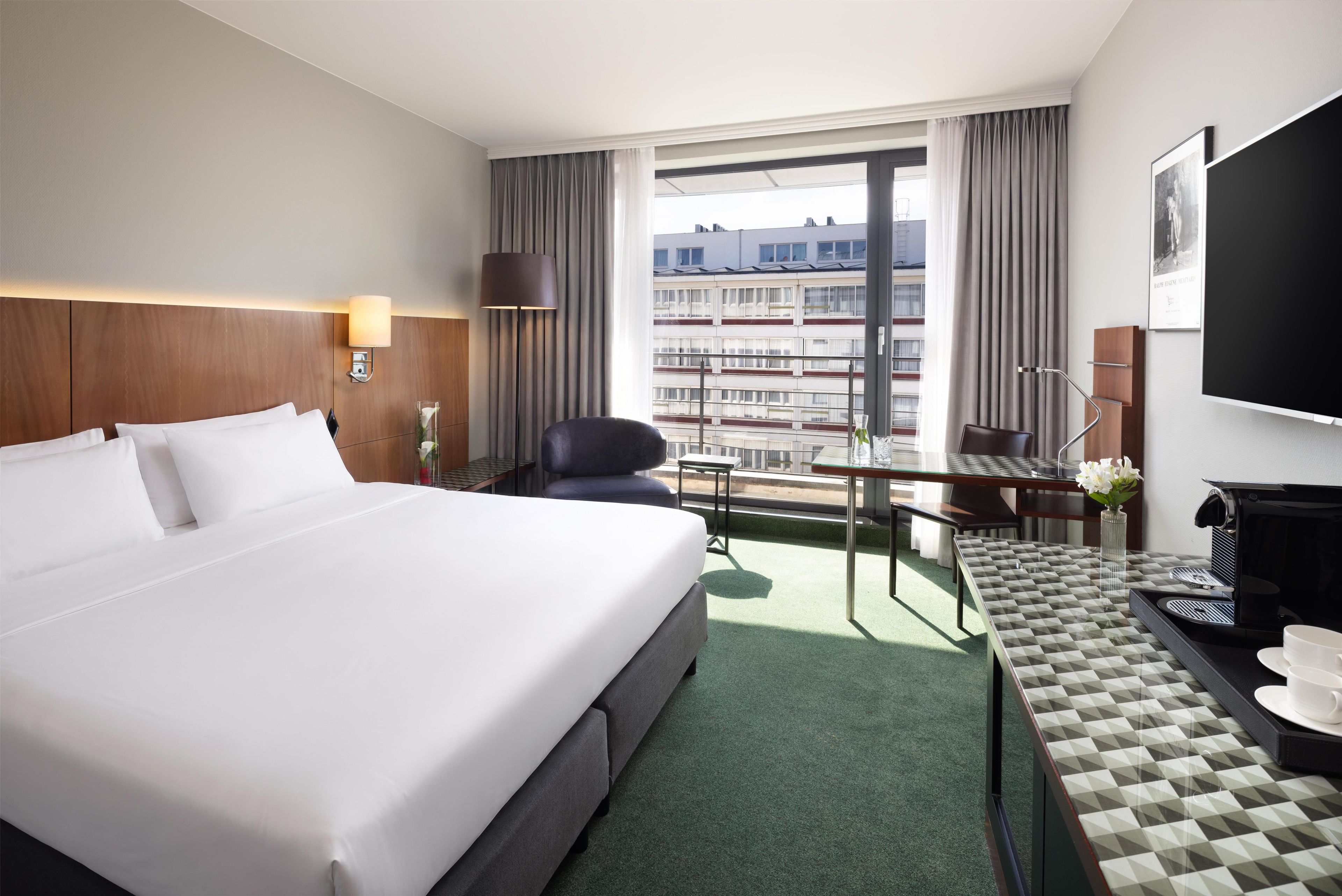 Quarto de hotel com uma cama grande, escrivaninha com cafeteira, cadeira, televisão e vista para edifícios pela janela.