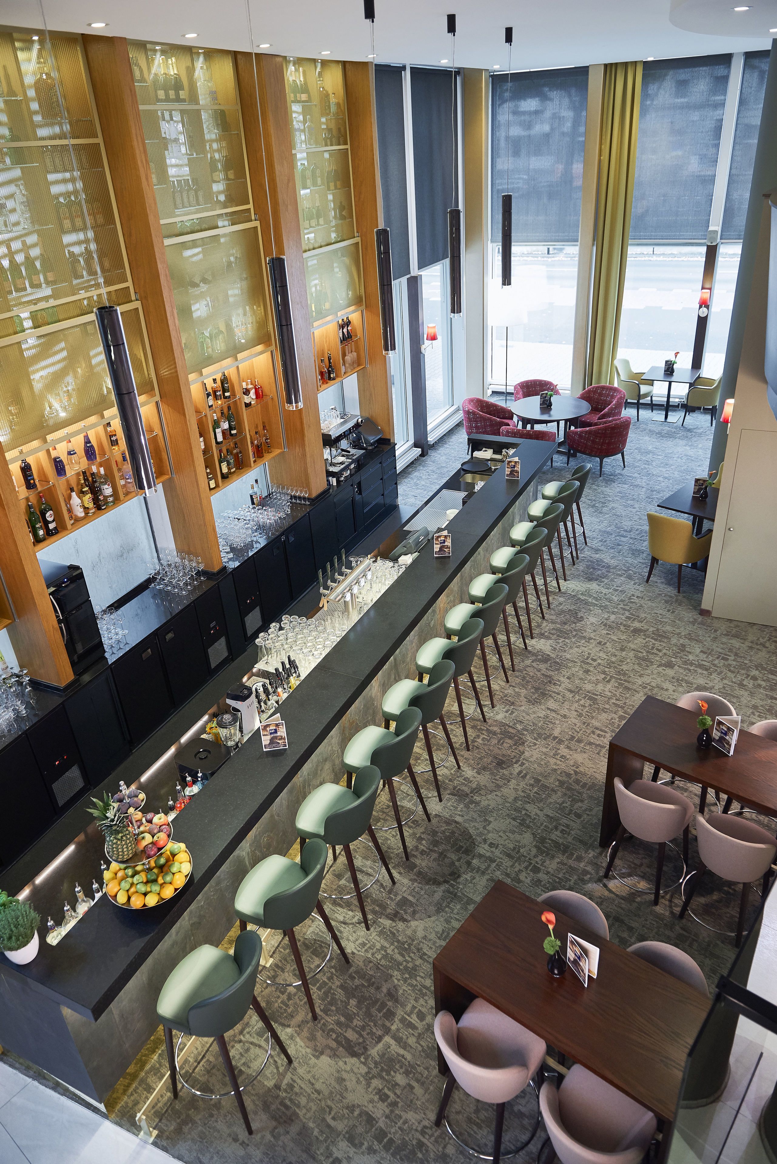 Uma área de bar com um balcão extenso, diversas opções de assento e vista da cidade através de amplas janelas.